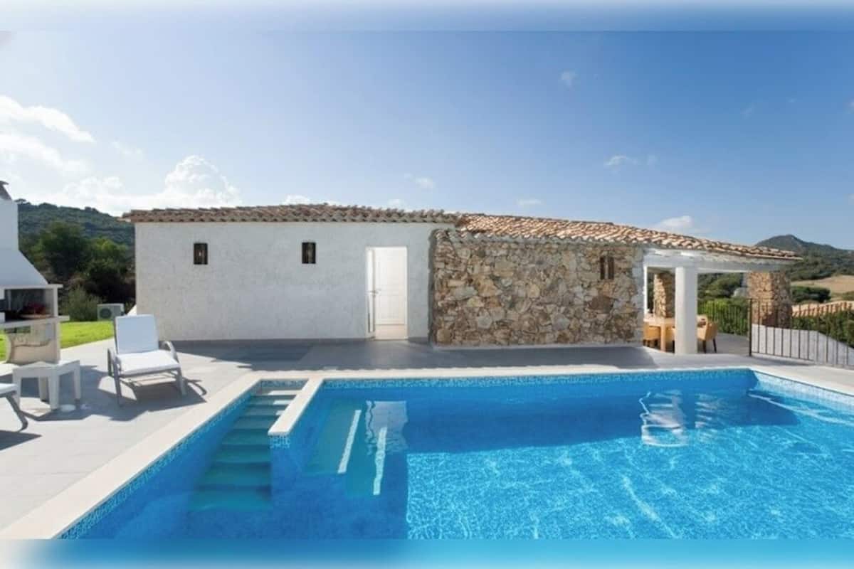 Uma área à beira da piscina com um muro de pedra, um edifício branco com uma porta e um céu azul claro.
