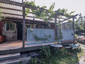 Um deck de madeira com pérgola, treliça branca decorativa e uma pequena mesa redonda com um banco.