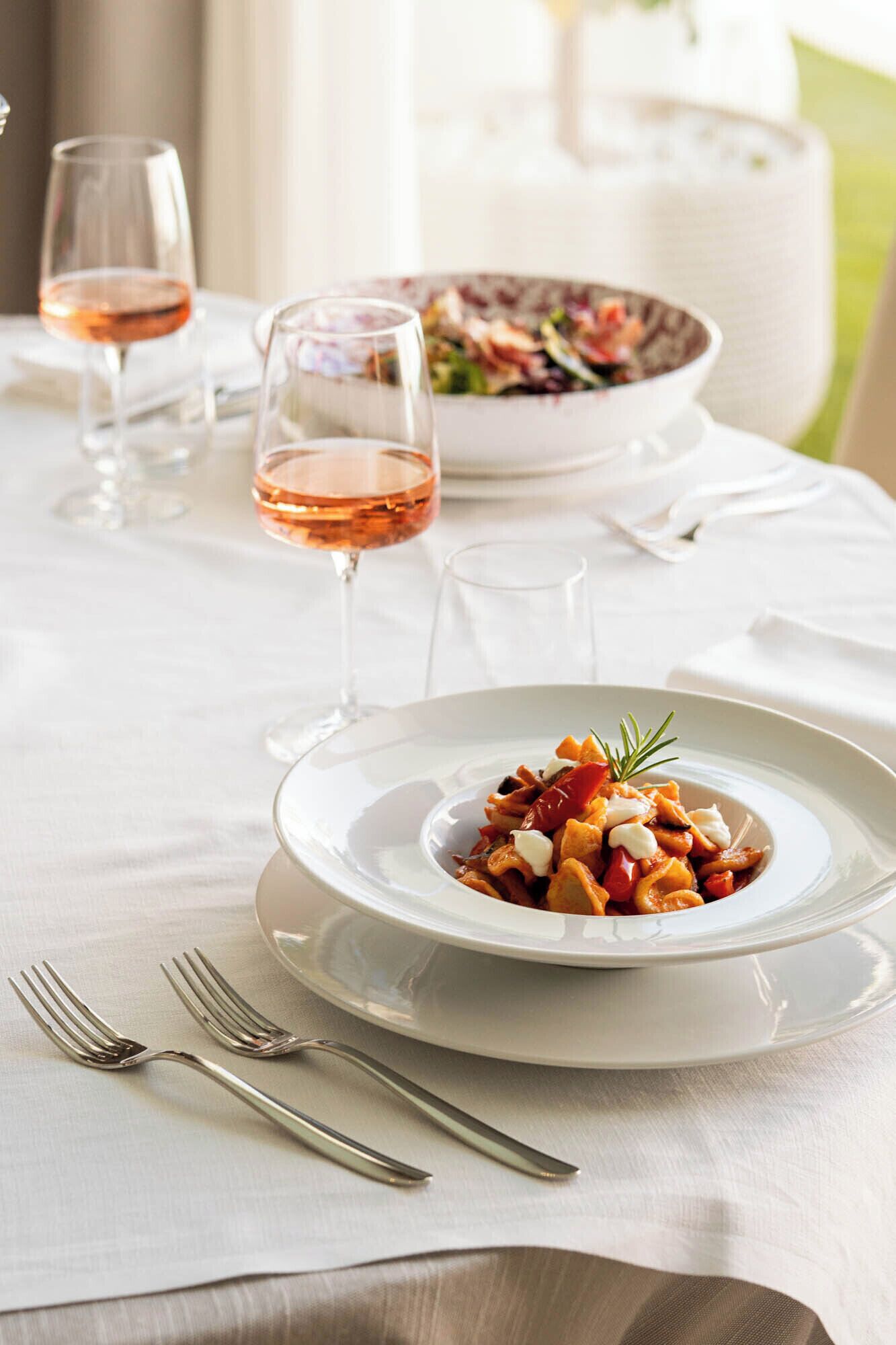 Uma mesa de jantar com um prato de comida, dois copos de vinho e uma saladeira ao fundo.