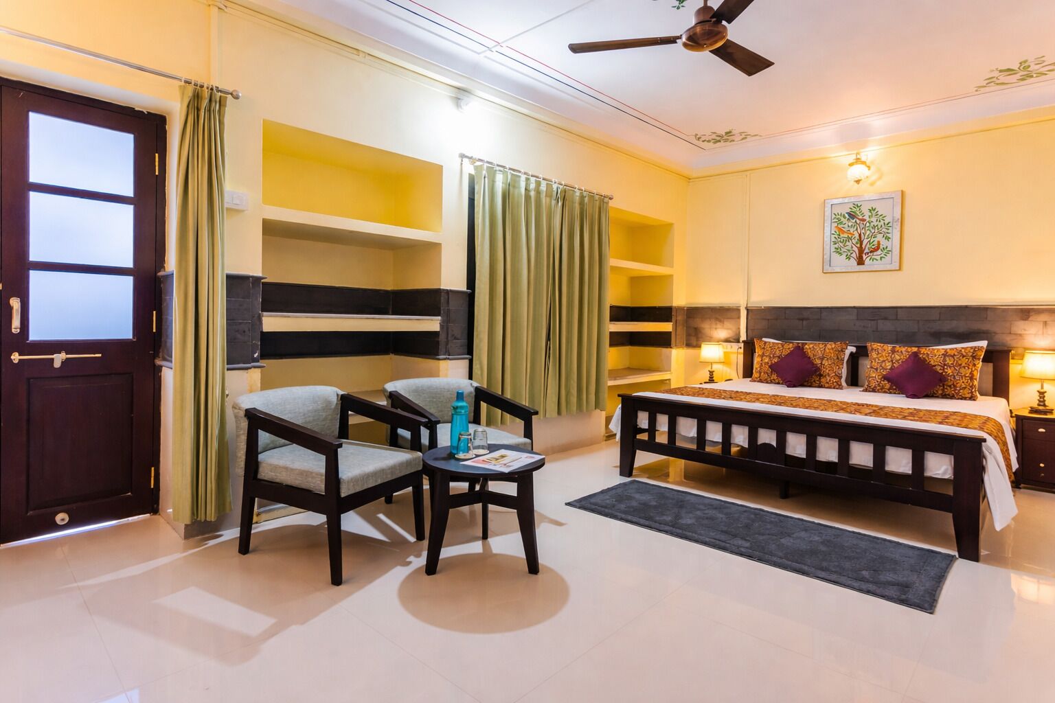 Quarto de hotel com cama, duas cadeiras, uma mesinha, ventoinha no teto e um quadro na parede.