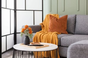 Uma sala de estar moderna com um sofá cinzento, uma manta amarela, almofada estampada, um vaso com flores laranjas e uma mesa de centro redonda com um controle remoto e uma bandeja.