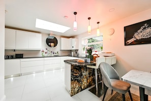 Uma cozinha moderna com armários brancos, uma ilha de mármore, luminárias pendentes e um espaço de jantar com mesa e cadeira.