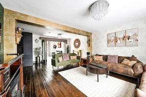 Uma sala de estar ampla com piso de madeira, um sofá verde, um sofá marrom, uma mesa de centro e um lustre.
