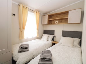 Quarto com duas camas, uma janela com cortinas, uma estante de madeira com uma decoração em formato de coração e uma mesa de cabeceira.