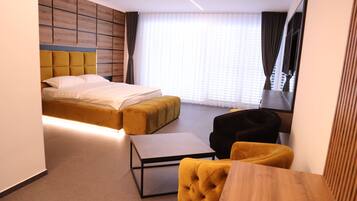 Quarto de hotel moderno com uma cama, uma mesinha, uma cadeira e uma janela grande com cortinas.