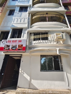 Um edifício de vários andares com a placa DSA Ruby Inn, apresentando uma série de janelas com grades de metal e uma unidade de ar condicionado visível.