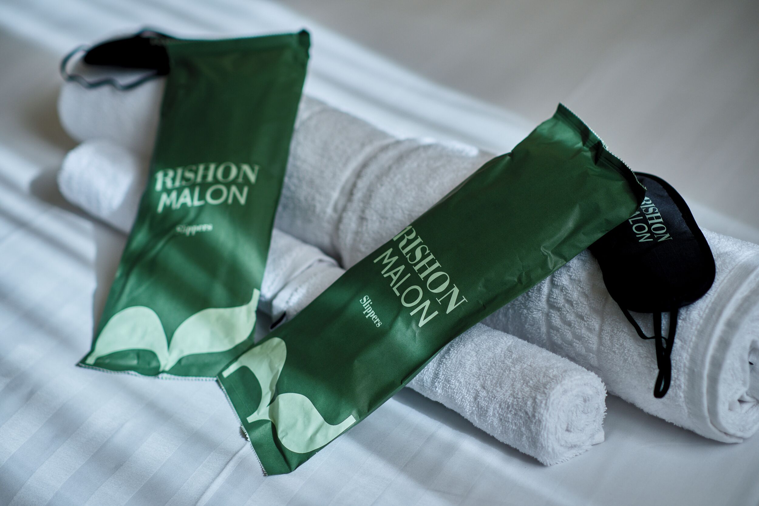 Toalhas brancas bem organizadas e um pacote verde com a inscrição 'RISHON MALON Chinelos' sobre a cama.