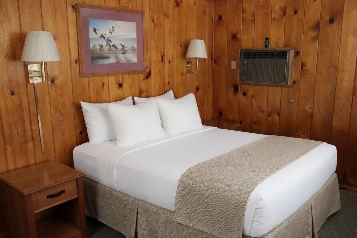 Quarto com painéis de madeira, uma cama, duas luminárias fixadas na parede, um quadro emoldurado e uma abertura de ventilação.