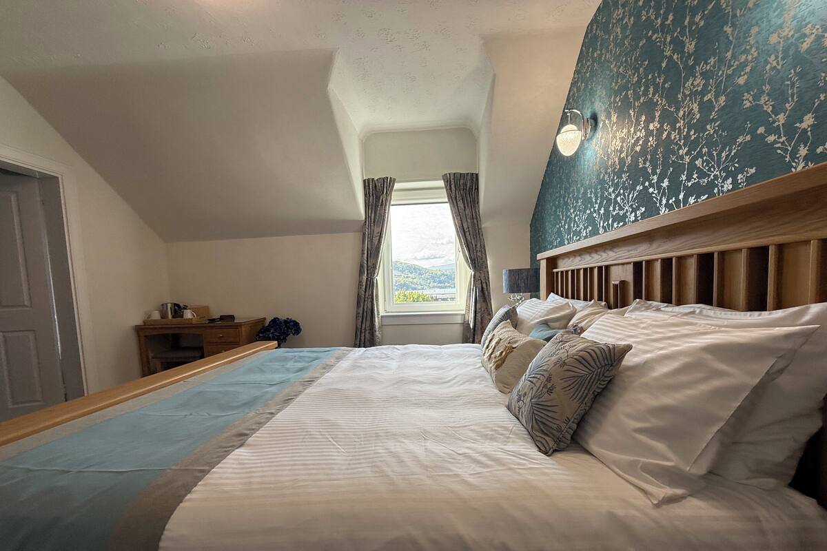 Quarto com uma cama grande, cabeceira de madeira, mesa de cabeceira, janela com cortinas e vista para o exterior.