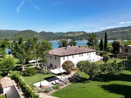 Um edifício branco com telhado em tijolos, rodeado por um jardim bem cuidado e vista para um lago e colinas.