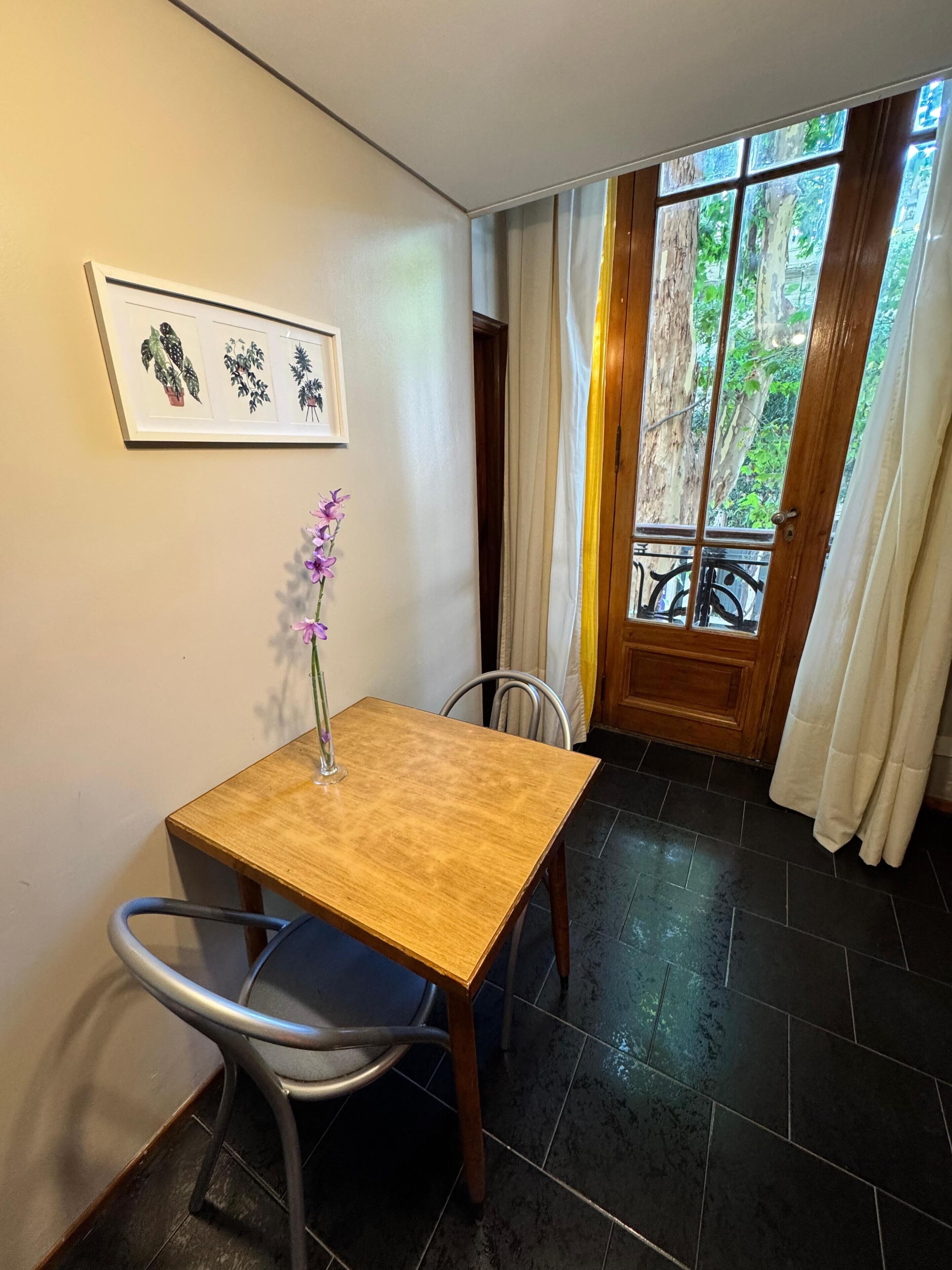 Uma sala de jantar com uma mesa de madeira, cadeiras de metal, um vaso com flores e gravuras botânicas emolduradas na parede.