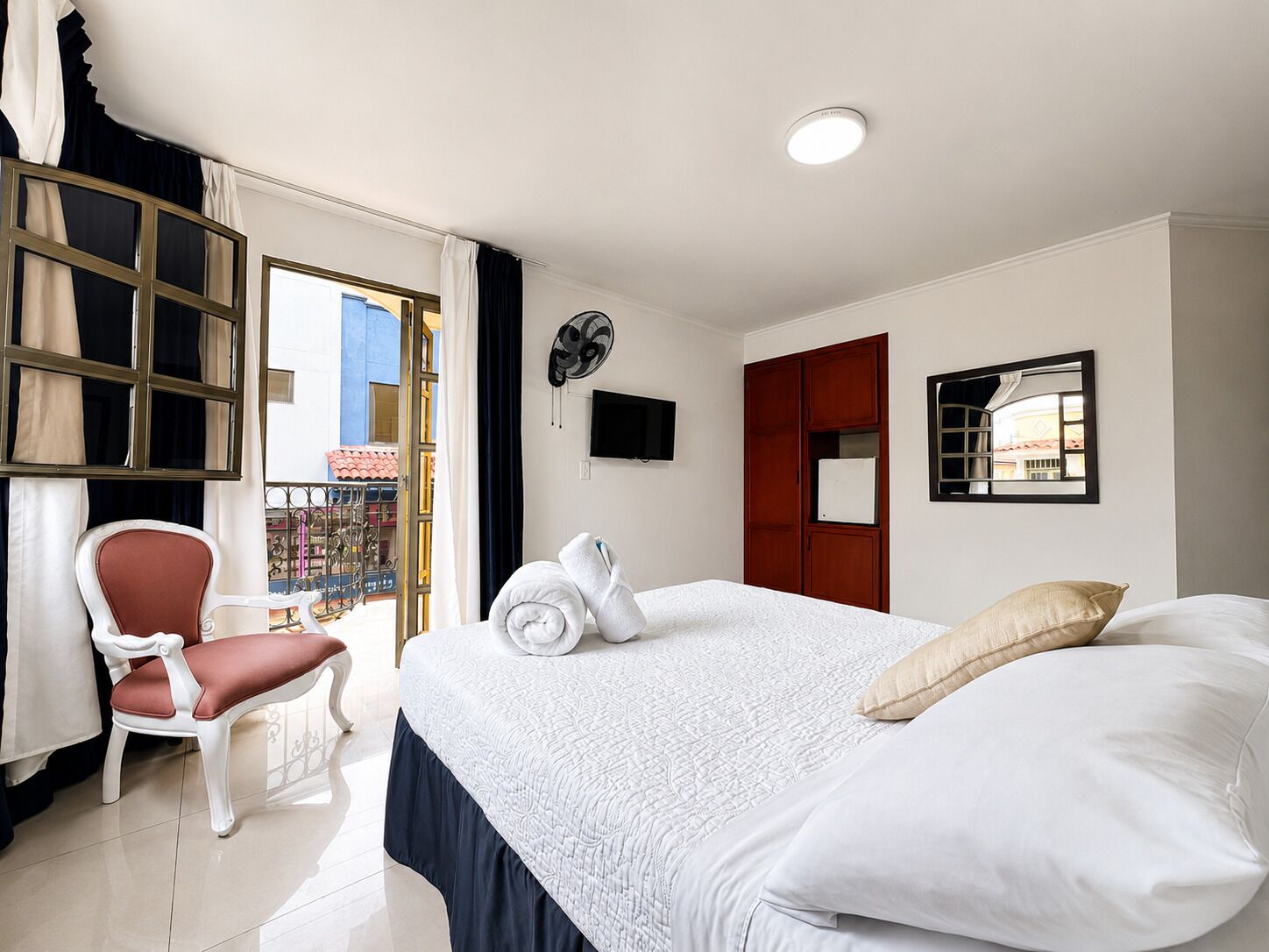 Quarto de hotel com cama, poltrona, televisão, espelho e janela com cortinas.