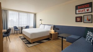 Quarto de hotel com cama, mesa pequena, cadeira, sofá e escrivaninha. Cortinas na janela e um quadro na parede.