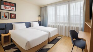 Quarto de hotel com duas camas, uma escrivaninha, uma cadeira, uma mesa de cabeceira, um abajur e uma janela com cortinas leves.