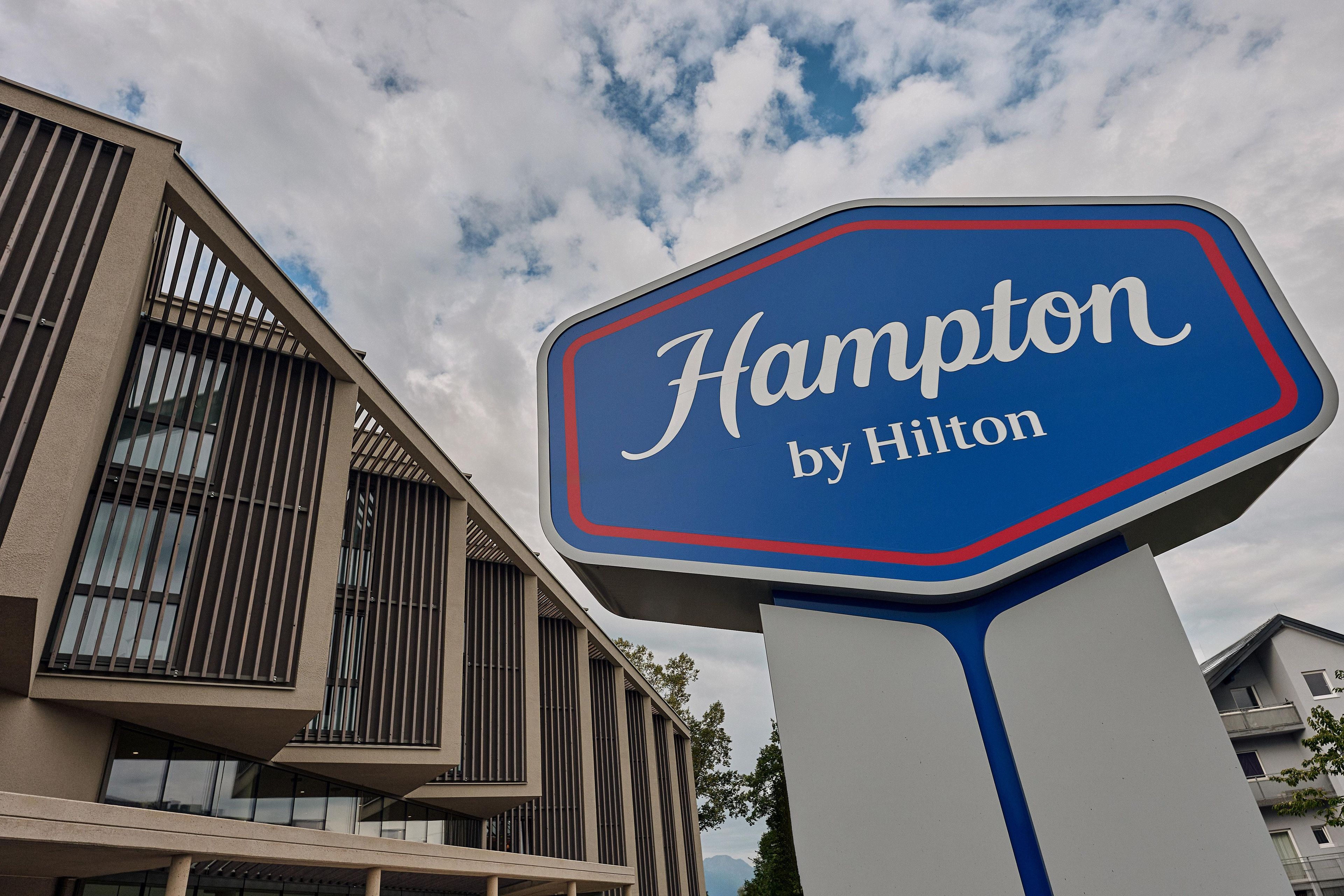 Placa de um hotel Hampton by Hilton, com um edifício moderno ao fundo.