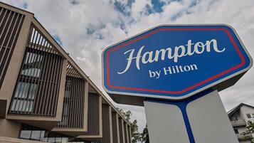 Placa de um hotel Hampton by Hilton, com um edifício moderno ao fundo.