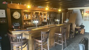 Uma área de bar com banquetas de madeira, um relógio e diversos garrafas e copos.