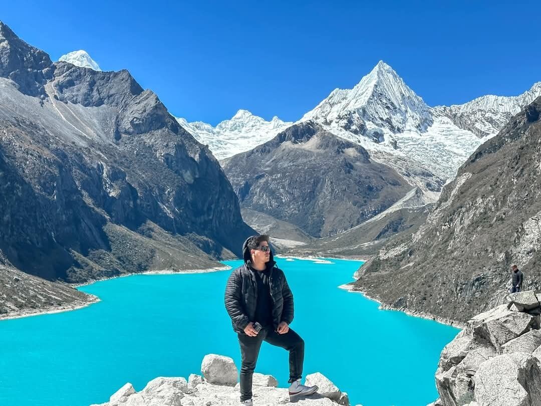 Uma pessoa sentada em pedras, com vista para um lago azul-turquesa e montanhas ao fundo.