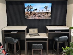 Um espaço de escritório moderno com uma televisão de tela grande fixada em uma parede escura, duas mesas com cadeiras e um laptop em uma delas.