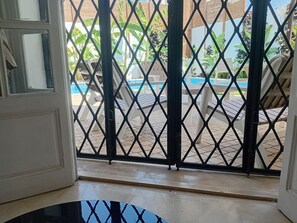 Visão através de um portão para uma área externa com piscina, espreguiçadeiras e plantas tropicais.