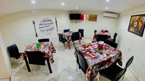 Uma área de refeições com mesas cobertas por toalhas temáticas de comida, cadeiras pretas e uma parede com um menu.