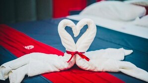 Uma cama com colcha vermelha e azul, uma toalha branca e uma toalha decorativa em forma de coração amarrada com um laço vermelho.