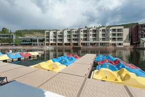 Um hotel na orla com um píer repleto de barcos a pedal colorido, um edifício moderno com varandas e um lago tranquilo ao fundo.