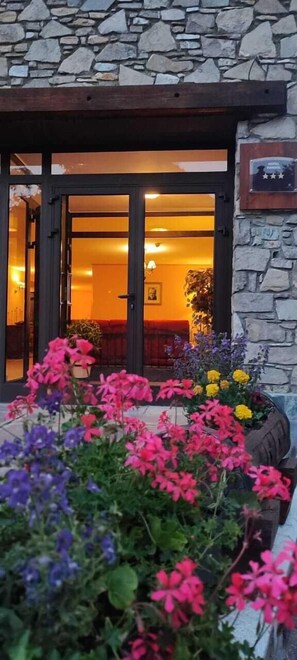 Fachada de pedra com porta de madeira, parede de pedra com uma placa e flores coloridas em primeiro plano.