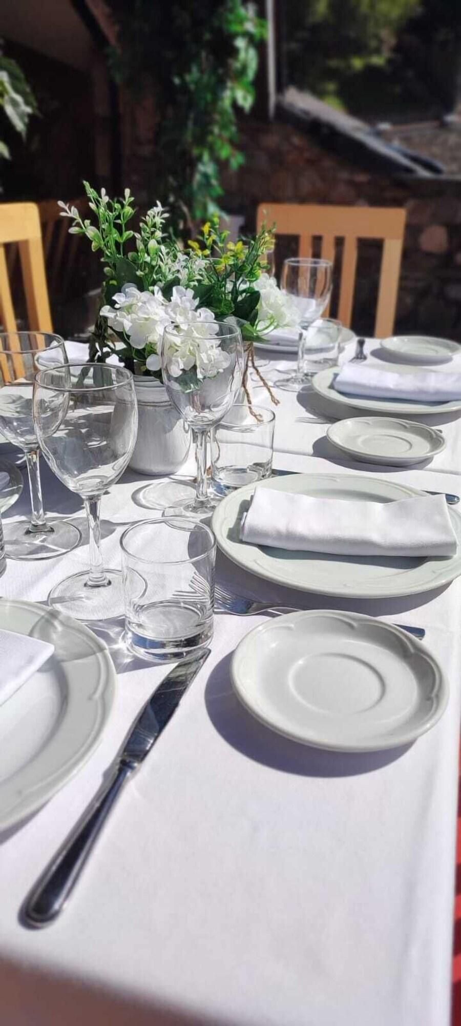 Uma mesa posta ao ar livre com toalhas brancas, copos de vidro transparente e um vaso com flores brancas.