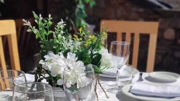 Uma mesa posta ao ar livre com toalhas brancas, copos de vidro transparente e um vaso com flores brancas.