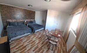 Quarto com duas camas, piso de madeira, mesa de jantar com cadeiras e janela com cortinas.