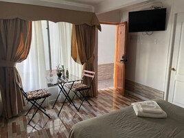 Um quarto com uma cama, uma mesa com cadeiras, uma televisão na parede e uma porta.