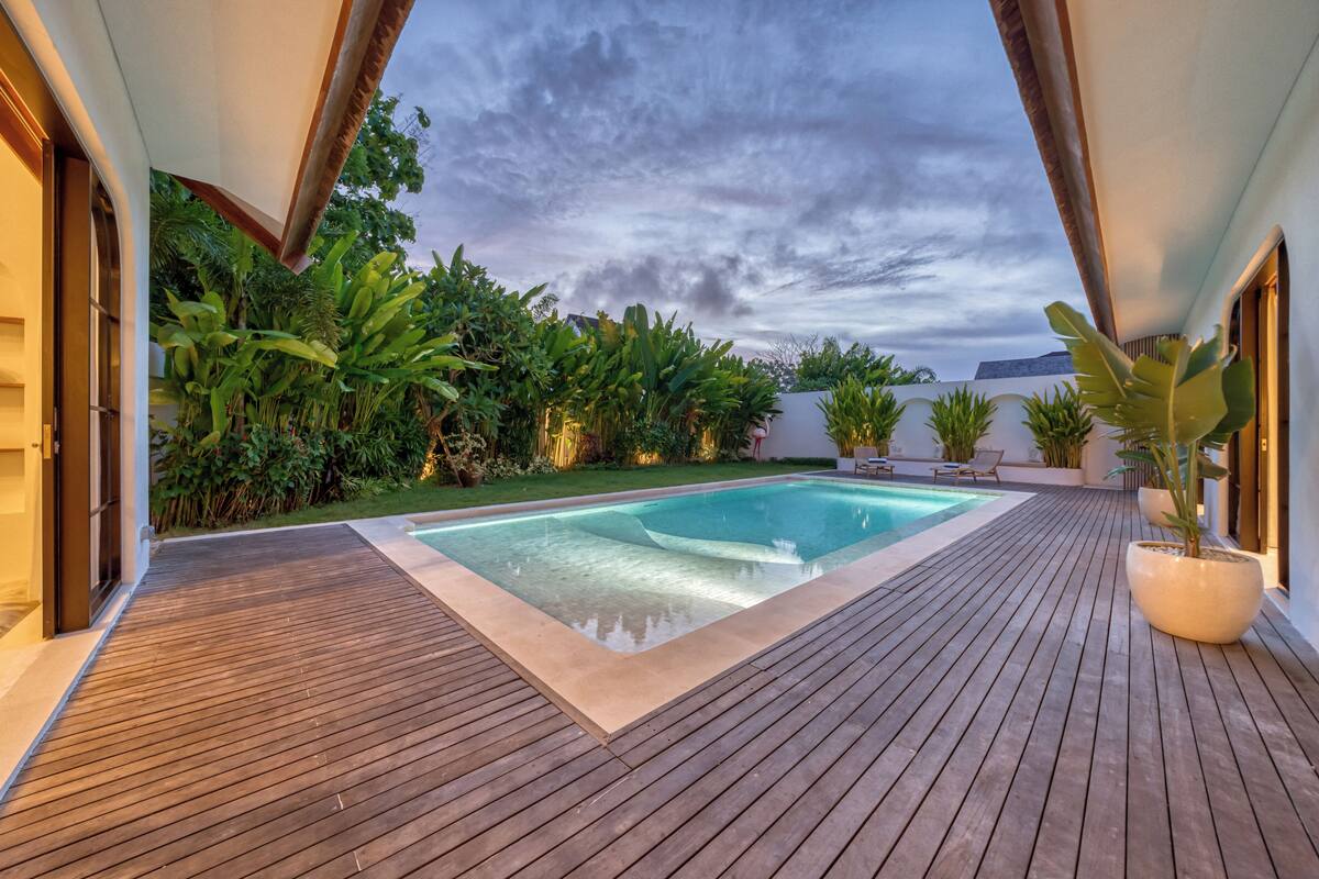 Piscina com deck de madeira, cercada por vegetação exuberante e um edifício moderno.