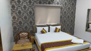 Quarto de hotel com uma cama, uma cadeira de madeira, uma mesinha e uma janela com persianas.