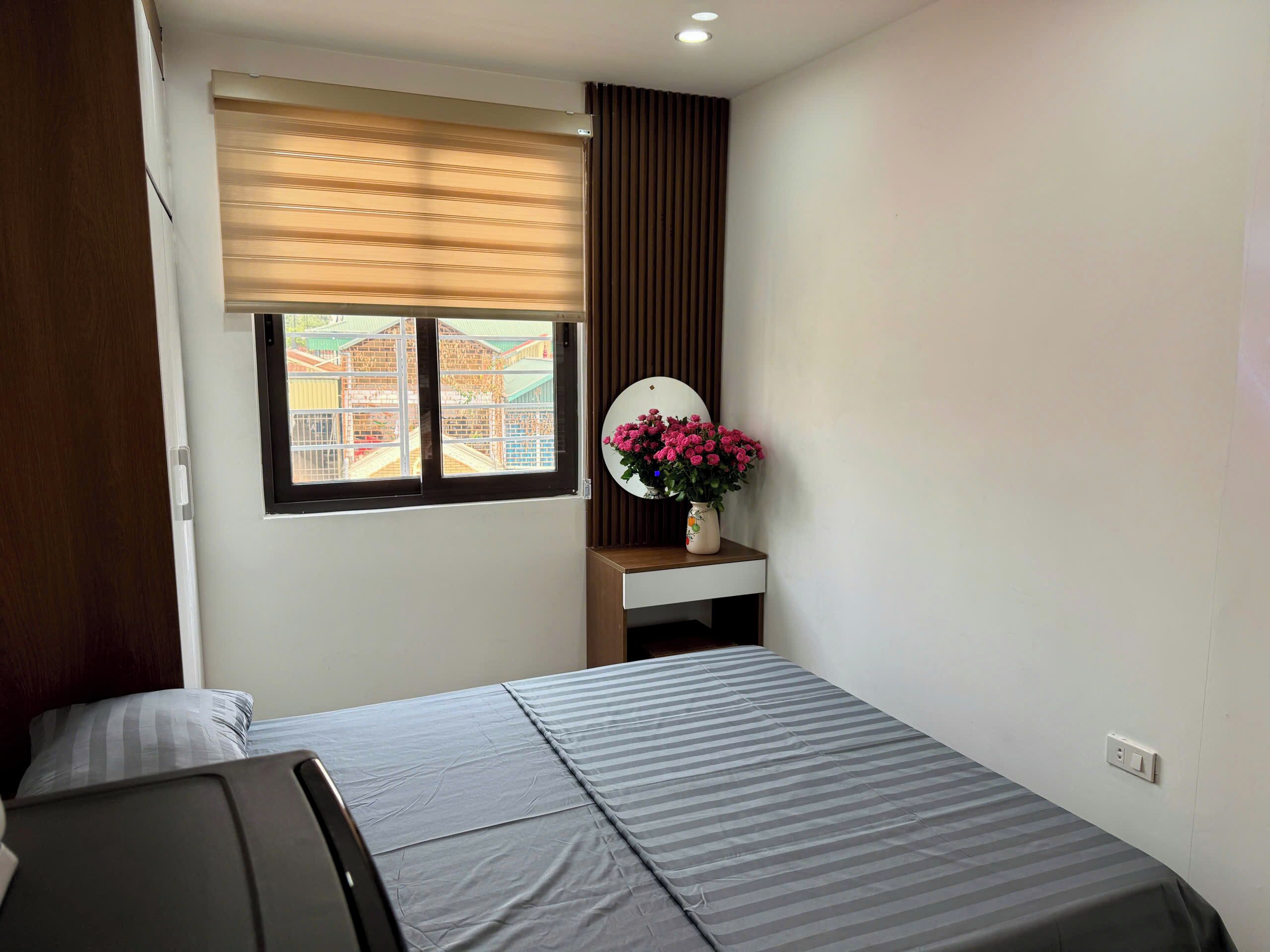 Um quarto com cama, janela com persianas, mesa de cabeceira com um vaso de flores e um espelho.