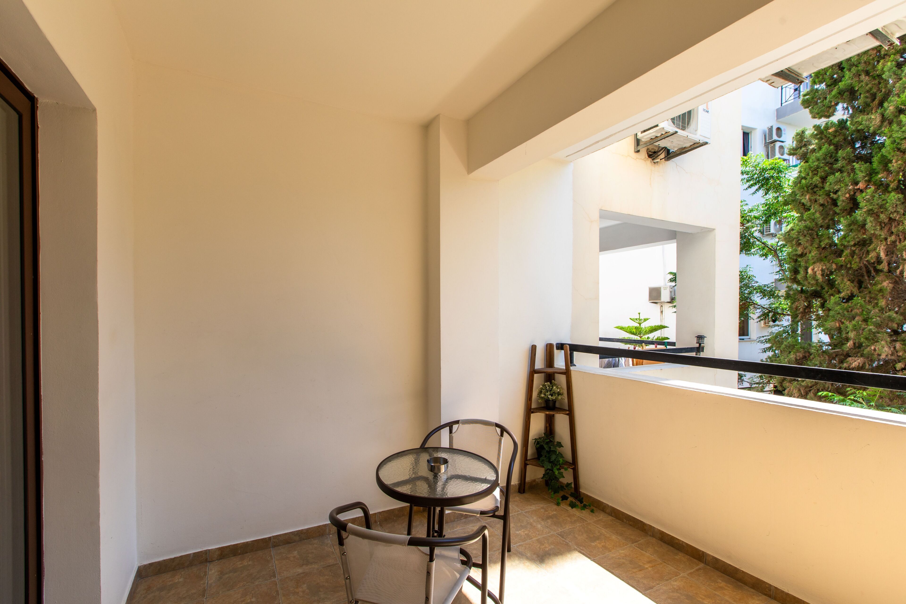 Varanda com mesa e cadeiras de vidro, uma escada e vista para um edifício e árvores.