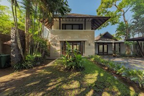 Uma casa de dois andares com parede de pedra, varanda coberta e um jardim com plantas tropicais.