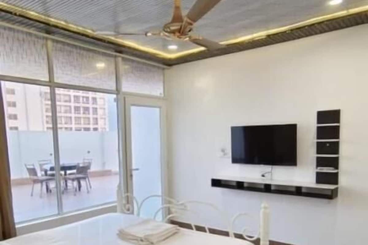 Quarto moderno com cama, ventilador de teto, TV fixada na parede e vista para o exterior.
