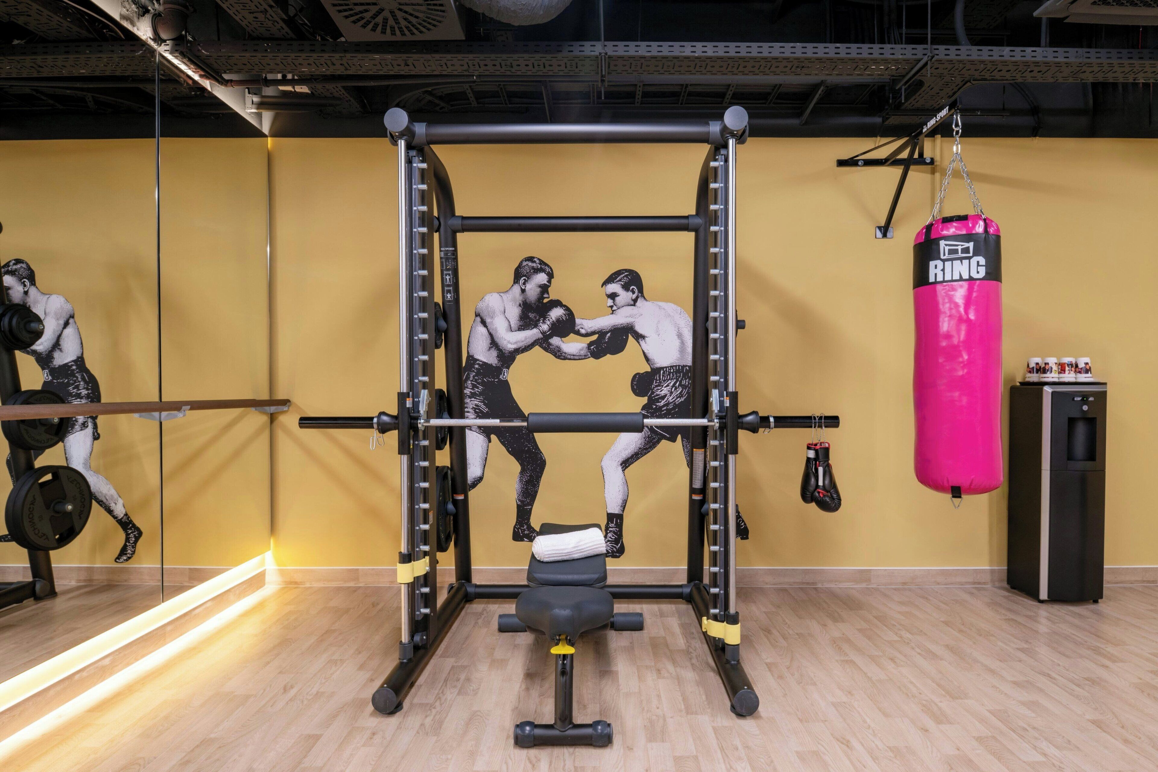 Uma academia com suporte para agachamento, saco de pancadas e um espelho refletindo o ambiente.