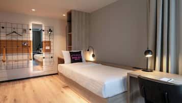 Quarto de hotel moderno com piso de madeira, cama com colchão branco e travesseiro com texto, escrivaninha com cadeira e espelho na parede.