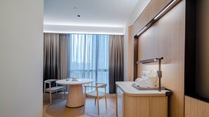 Quarto de hotel moderno com mesa de jantar redonda, duas cadeiras, pequena cozinha com cafeteira e uma janela grande com cortinas.