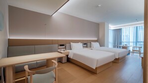 Quarto de hotel moderno com uma cama grande, uma escrivaninha, uma cadeira e vista para a cidade através de janelas amplas.