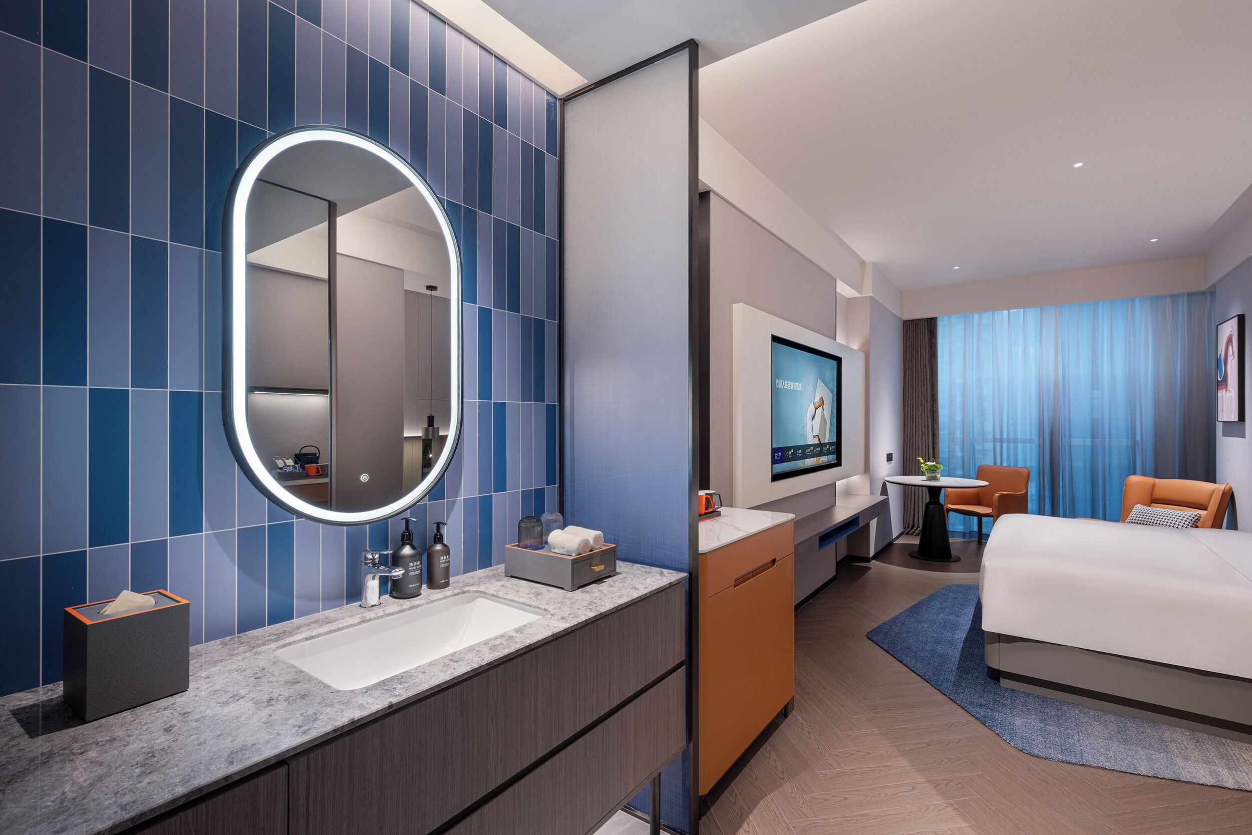 Quarto de hotel moderno com um espelho grande, televisão, cama, secretária e casa de banho com lavatório e espelho.