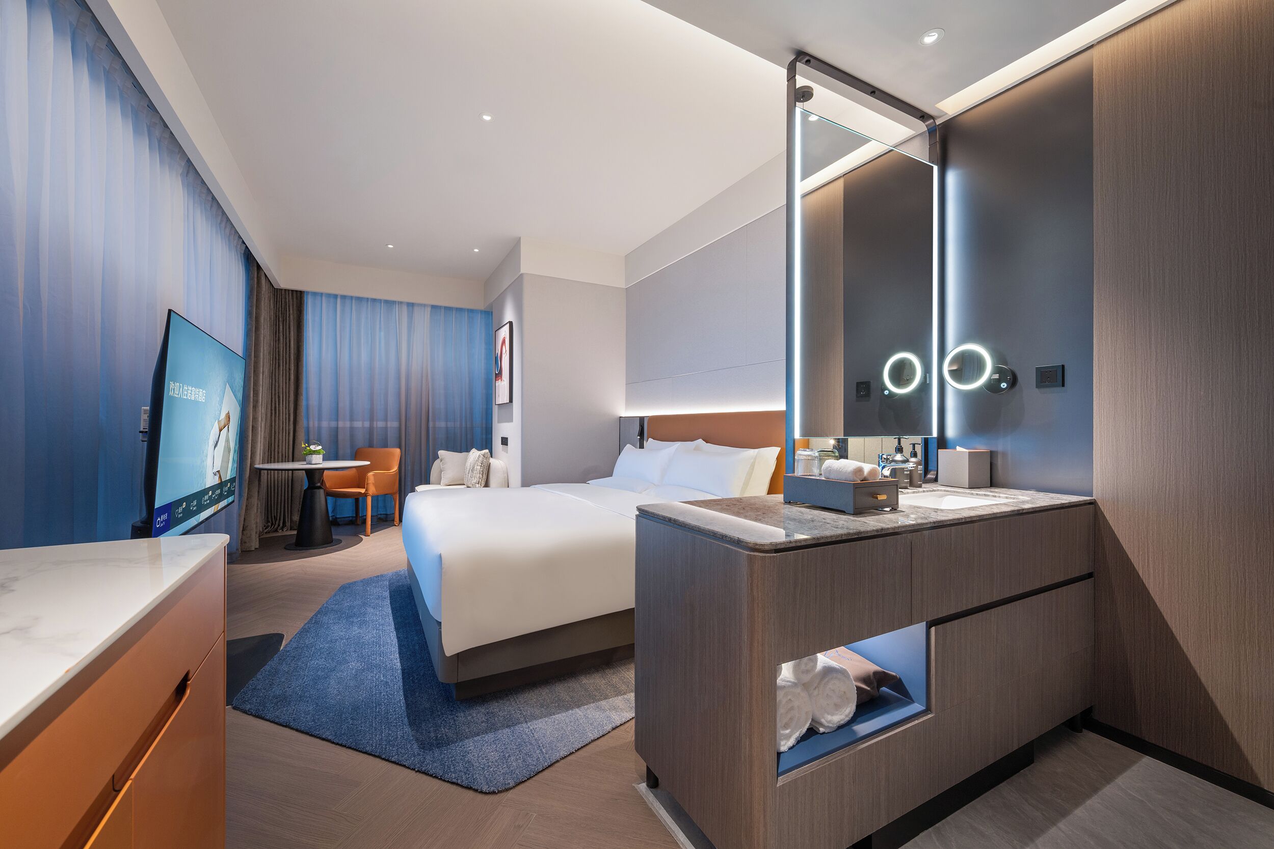 Quarto de hotel moderno com cama, escrivaninha, cadeira, banheiro com espelho e televisão.