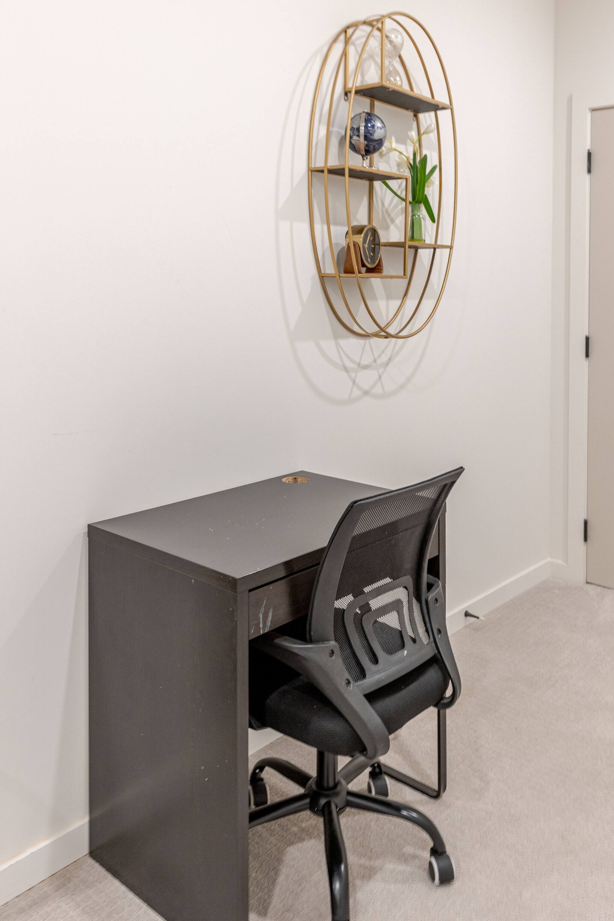 Uma mesa de escritório moderna com uma cadeira, uma prateleira suspensa dourada com objetos decorativos, e uma porta ao fundo.