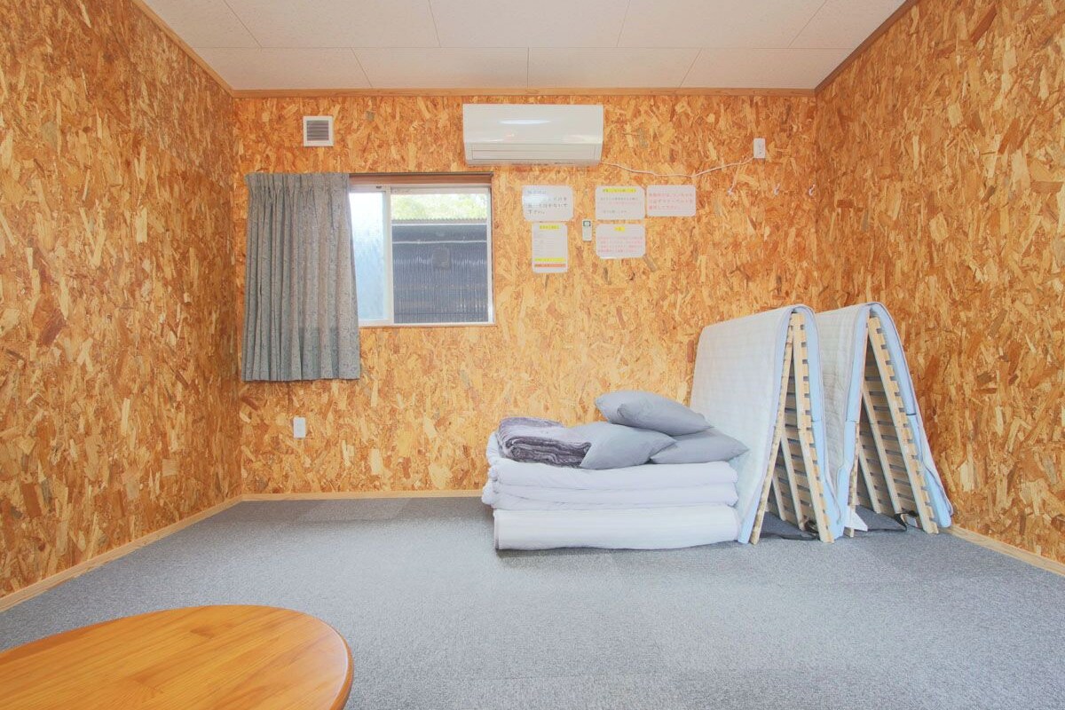Quarto com parede de madeira, janela com cortinas, cama com almofadas, mesa e aparelho de ar condicionado.