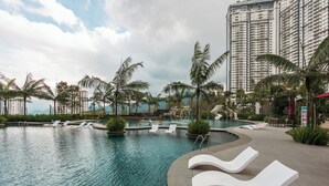 Uma piscina com espreguiçadeiras, palmeiras e edifícios altos ao fundo.
