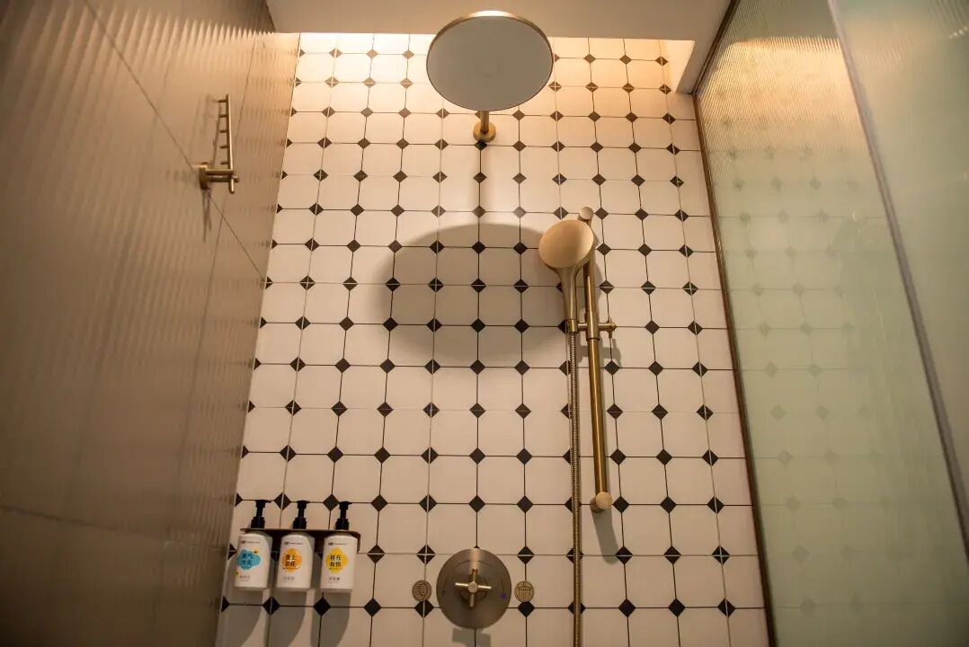 Um banheiro com parede de azulejos estampados geometricamente, chuveiro manual, espelho fixado na parede e um conjunto de produtos de higiene pessoal.