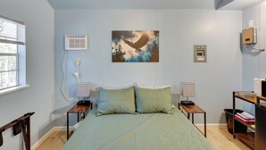 Um quarto com cama, duas mesas de cabeceira, ar-condicionado fixado na parede, quadro de eletricidade na parede e uma janela com persianas.