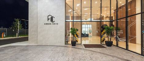 Entrada moderna de hotel com fachada de vidro, logotipo na parede e plantas em vasos.